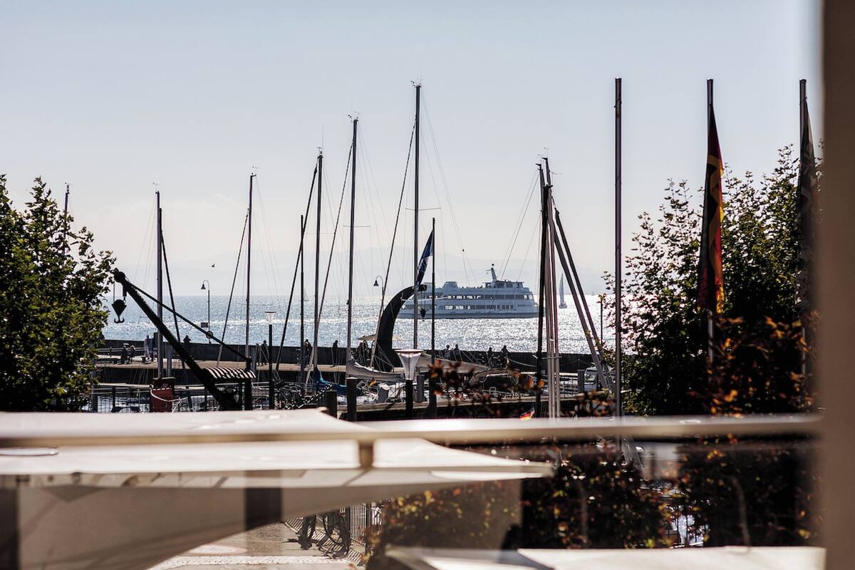 Marina com vários veleiros atracados, um grande navio de cruzeiro ao fundo e vista para a água.