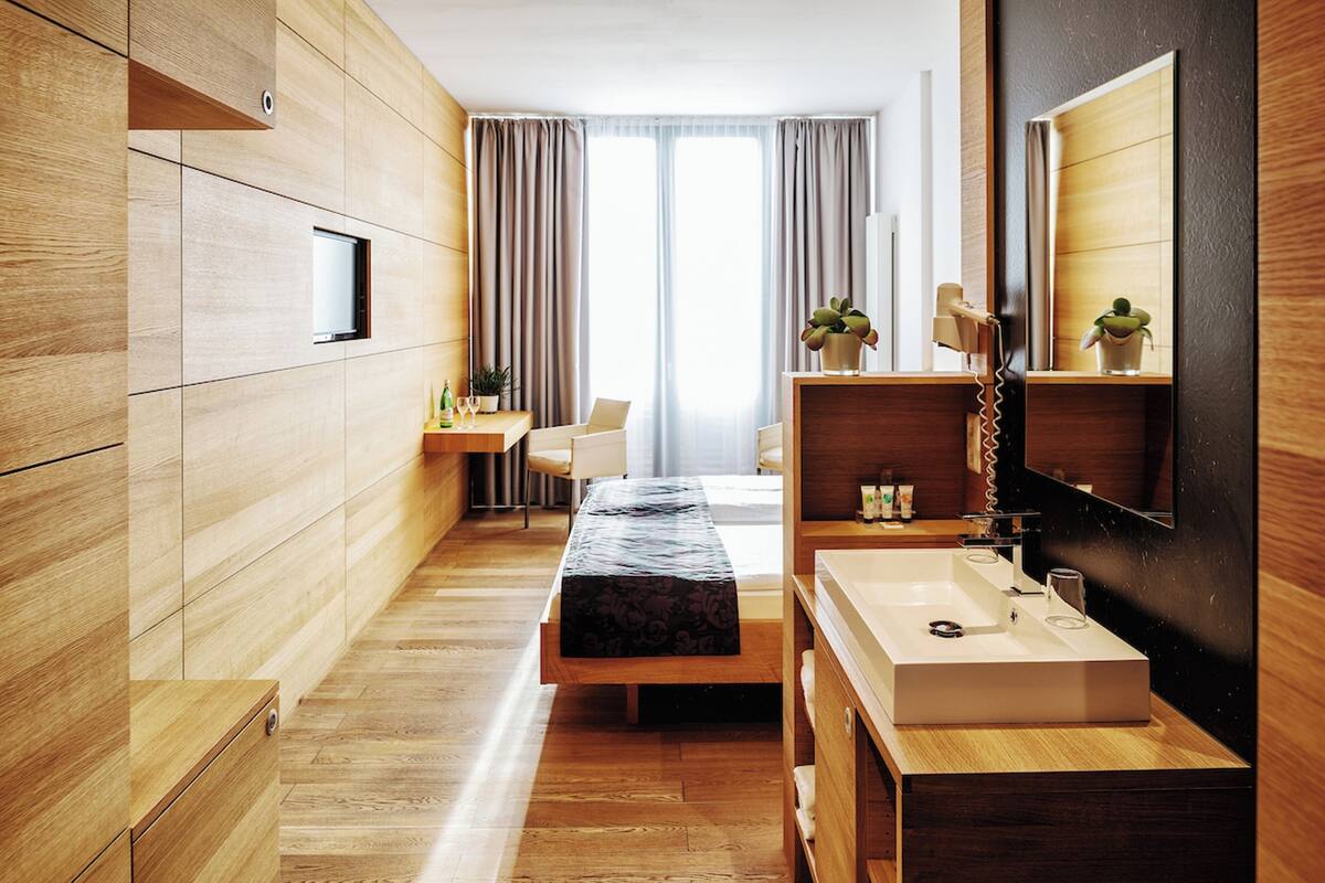 Quarto de hotel moderno com chão de madeira, cama com colcha estampada escura, pia branca, espelho grande e pequena mesa com uma planta.