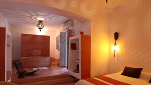 Quarto com cama, cadeira, mesa pequena, prateleira e banheiro com banheira e chuveiro.