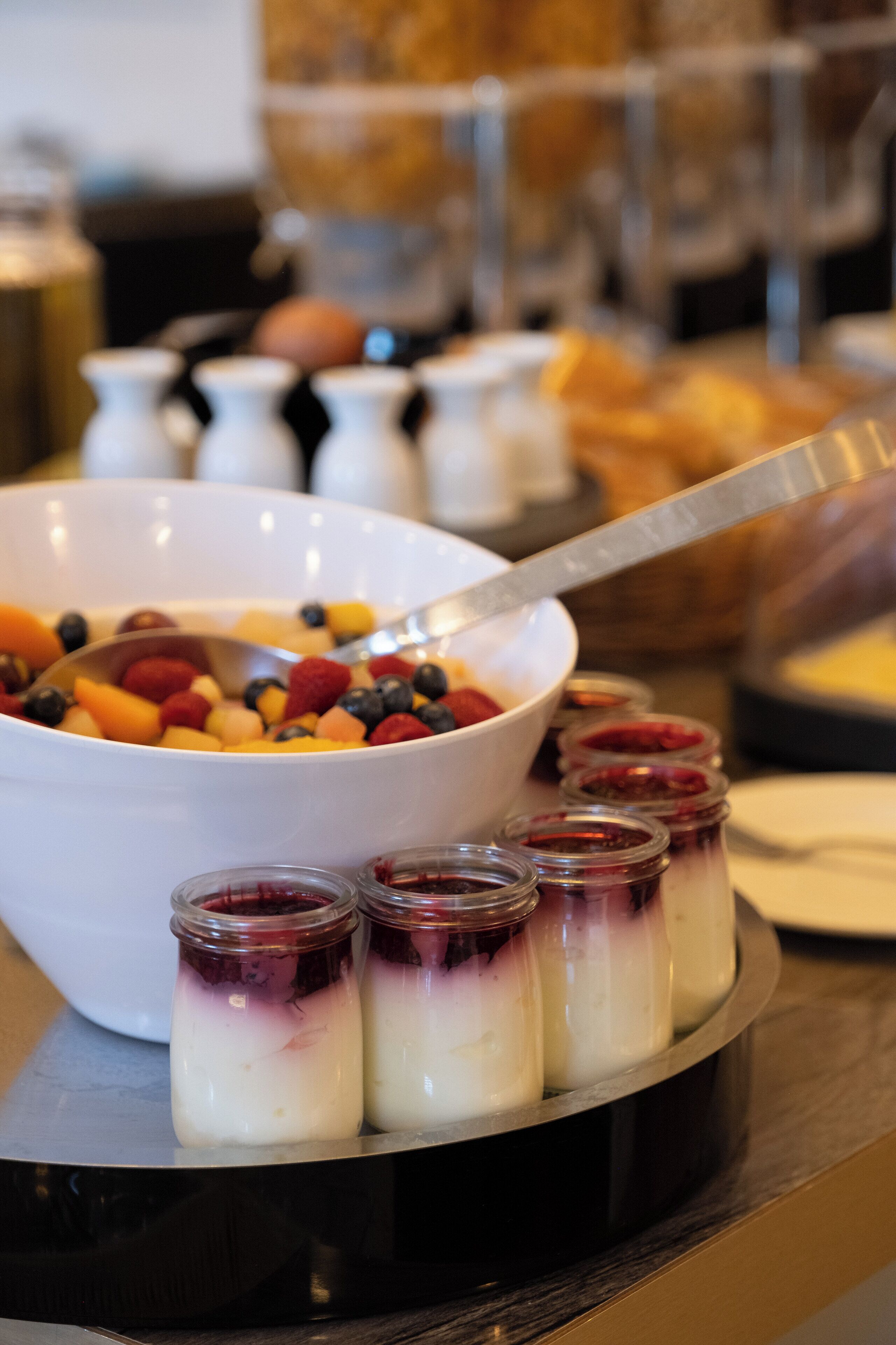 Um buffet de café da manhã com uma tigela de frutas variadas, uma colher e pequenos potes de geleia.