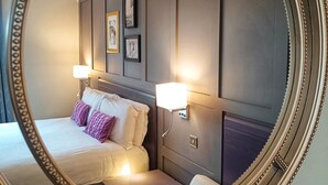 Quarto de hotel com cama grande, duas luminárias de cabeceira, TV na parede e quadros decorativos.