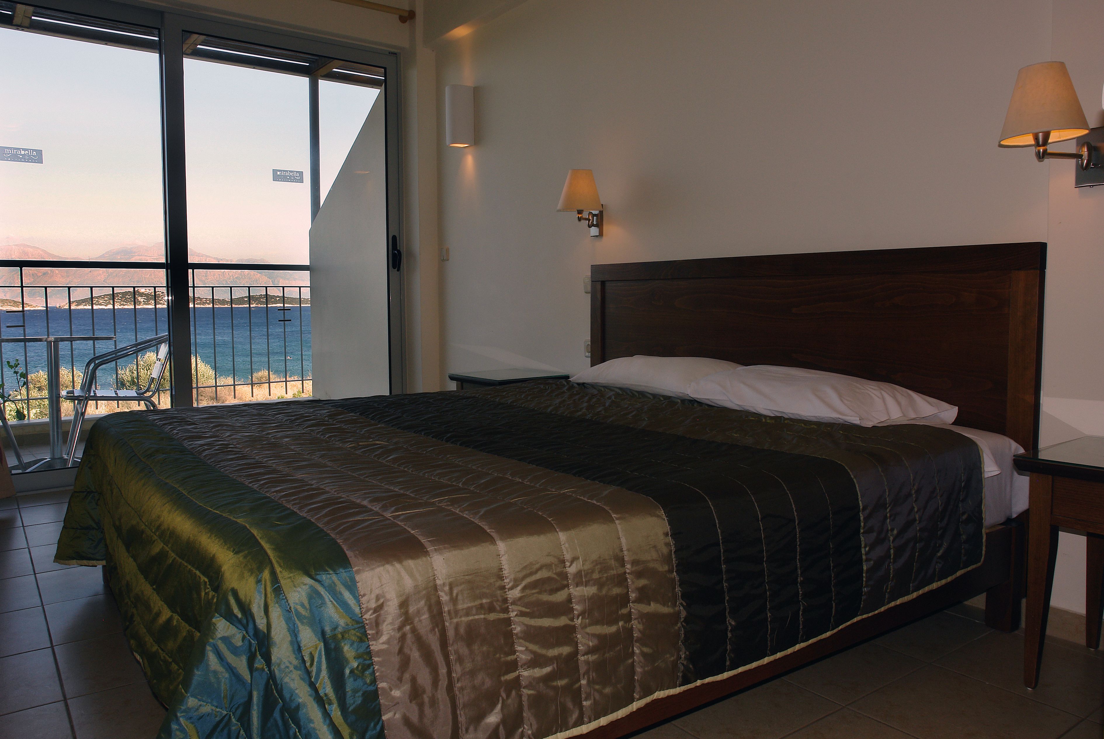 Quarto de hotel com uma cama grande, varanda com vista para o mar e duas luminárias fixadas na parede.