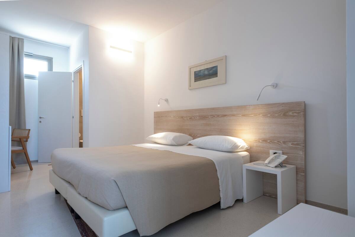 Quarto moderno com uma cama grande, mesas de cabeceira, um criado-mudo com telefone e um quadro na parede.