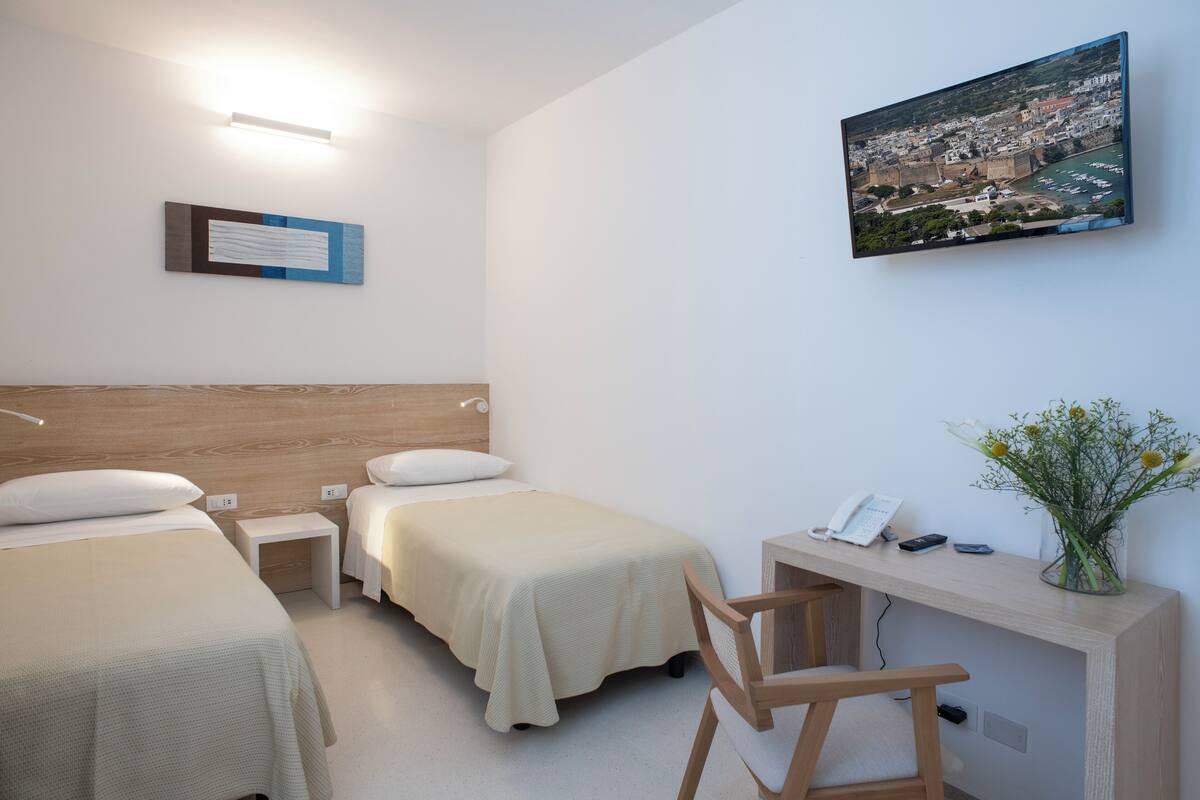 Quarto de hotel com duas camas, uma escrivaninha, uma cadeira, um telefone e um vaso com flores.