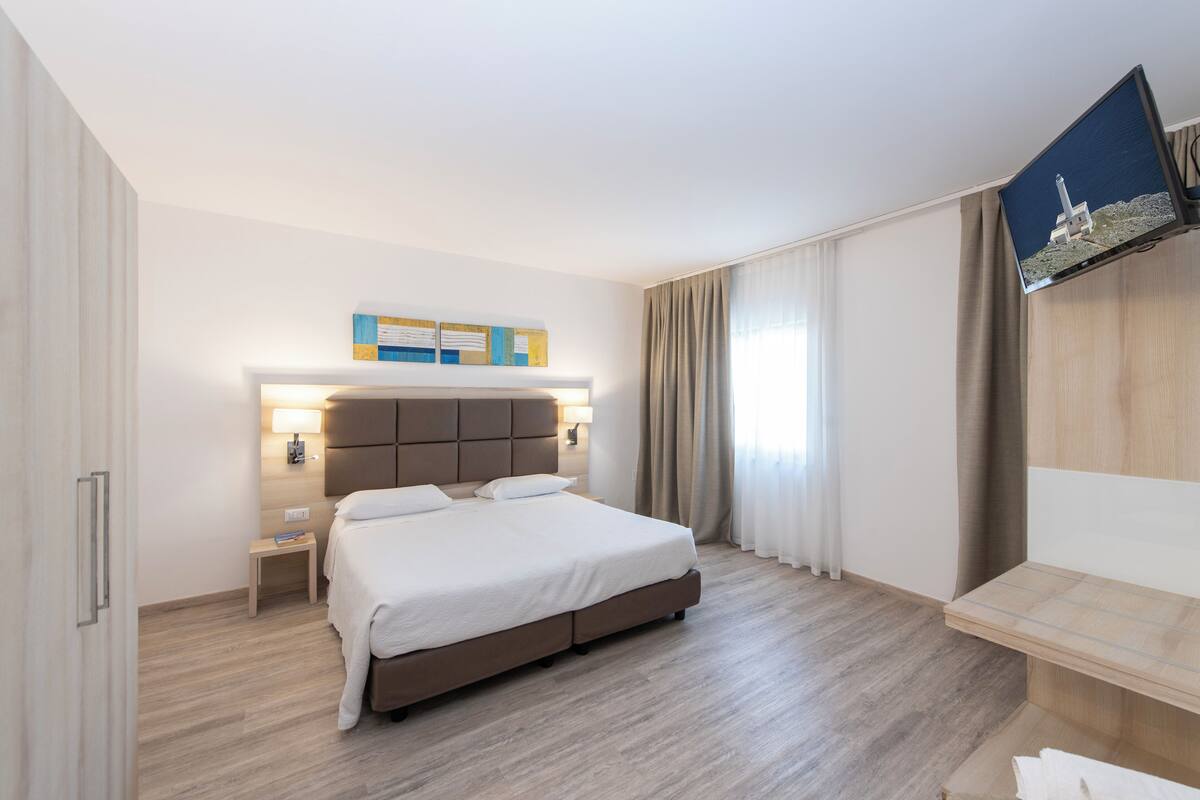 Quarto de hotel com uma cama grande, televisão de tela plana na parede, mesa de cabeceira com abajur e uma mesinha ao lado da cama.