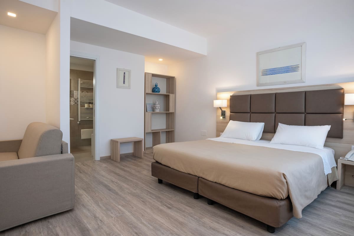Quarto de hotel moderno com uma cama grande, um sofá, uma mesa de cabeceira e um criado-mudo. Há uma porta que dá para um banheiro e um guarda-roupa.