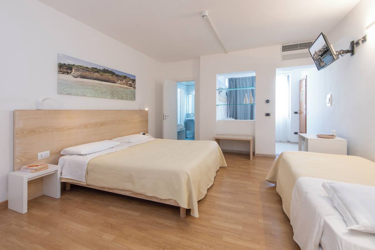 Quarto de hotel com duas camas, cabeceira de madeira, mesa de cabeceira, pequena escrivaninha, janela com cortinas e televisão na parede.