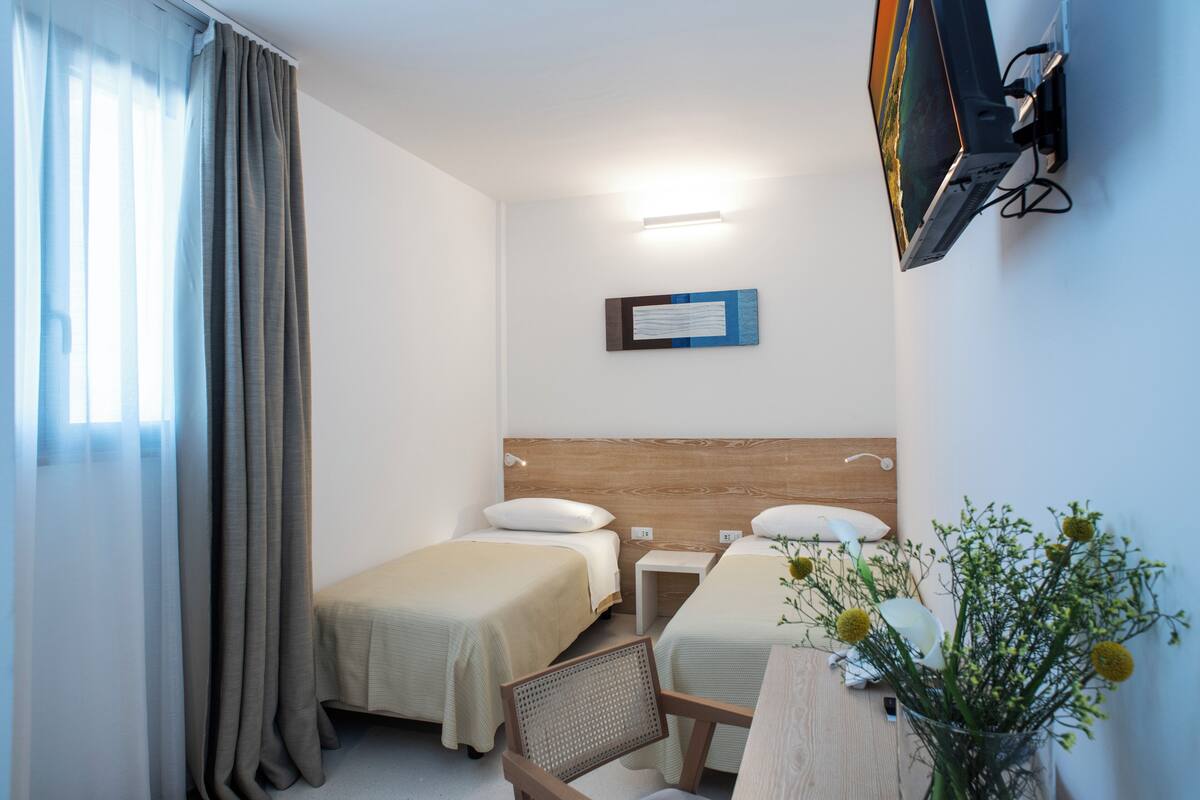 Quarto de hotel com duas camas de solteiro, uma escrivaninha com cadeira, televisão de tela plana na parede e uma janela com cortinas.