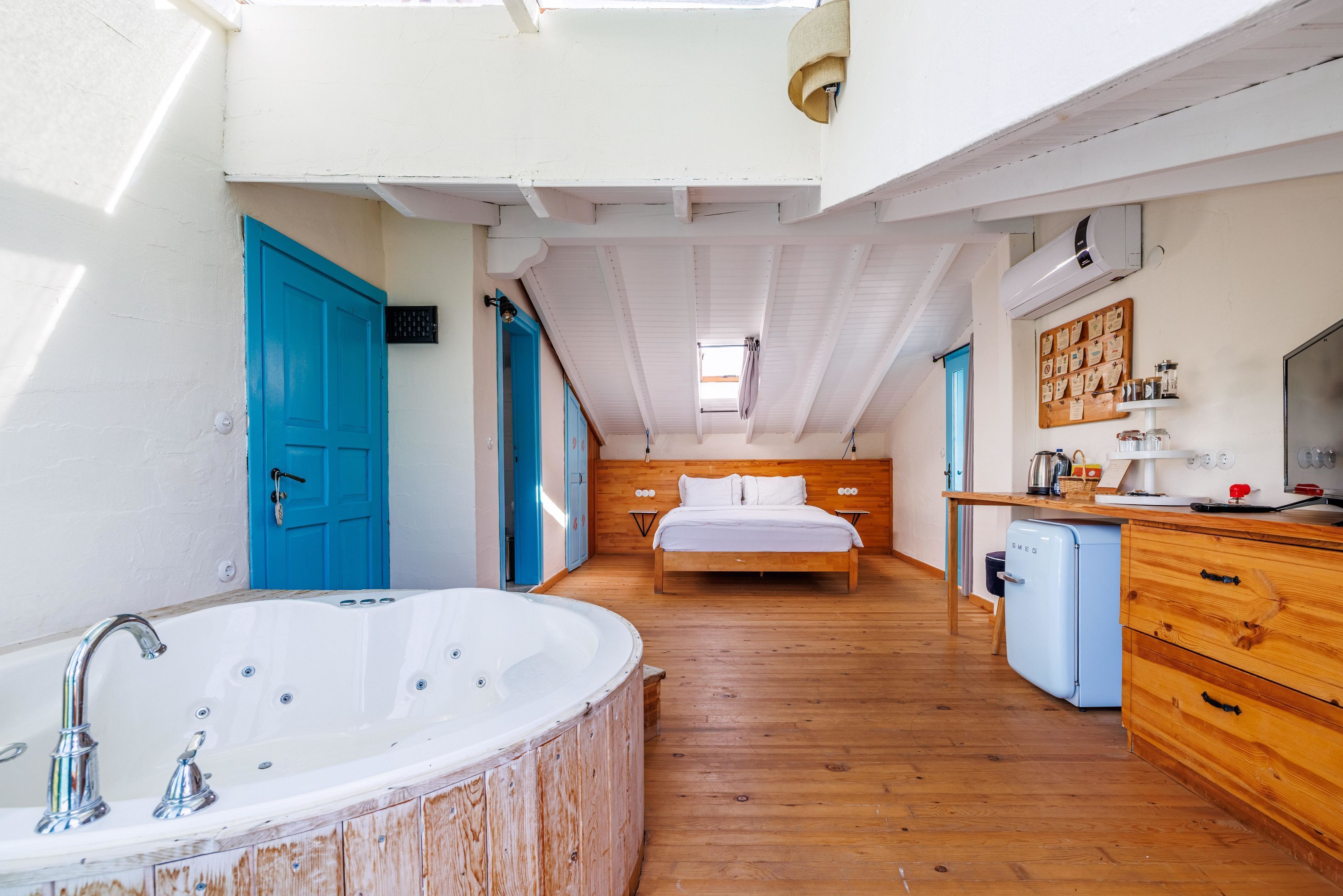 Quarto com piso de madeira, uma banheira grande, cama com roupa de cama branca, cômoda de madeira e uma pequena geladeira.