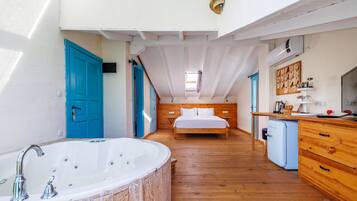 Quarto com piso de madeira, uma banheira grande, cama com roupa de cama branca, cômoda de madeira e uma pequena geladeira.