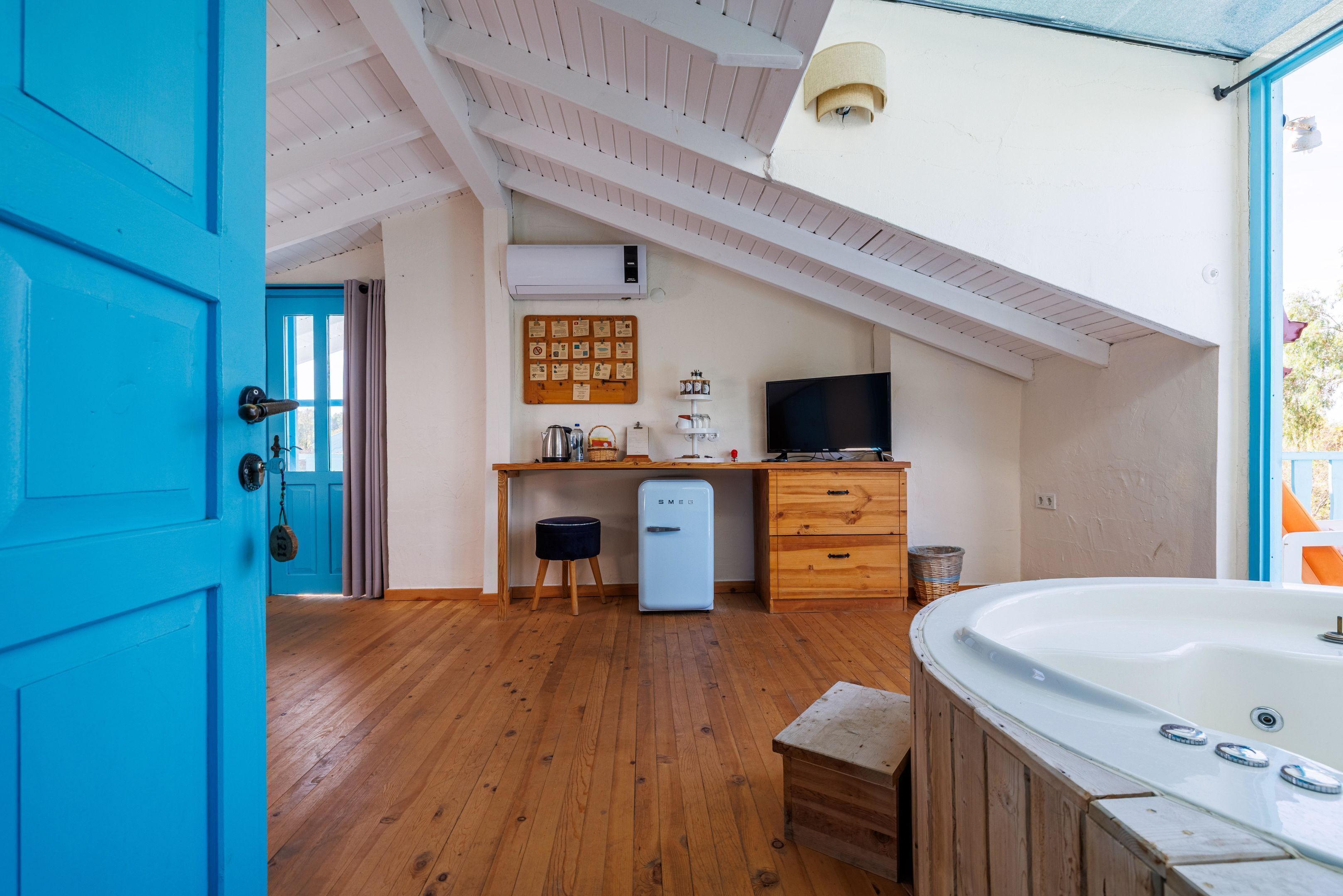 Quarto com piso de madeira, porta azul, banheira branca, escrivaninha de madeira, televisão e um pequeno frigorífico.