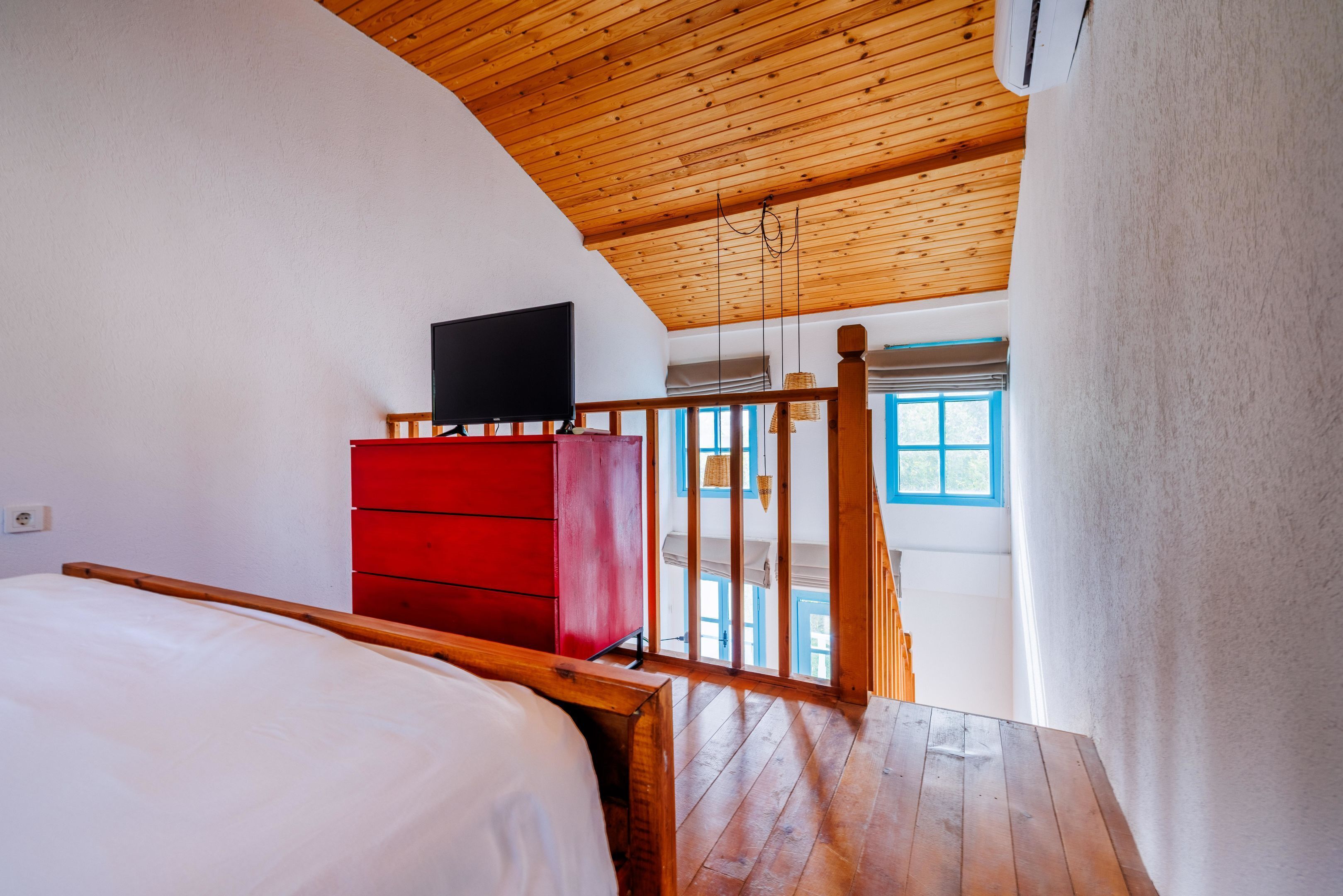 Quarto com teto de madeira, uma cama, penteadeira vermelha, televisão e varanda com vista.