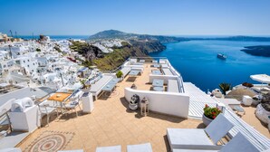 Terraço no telhado com móveis brancos, piso estampado e vista para o mar e uma cidade costeira.