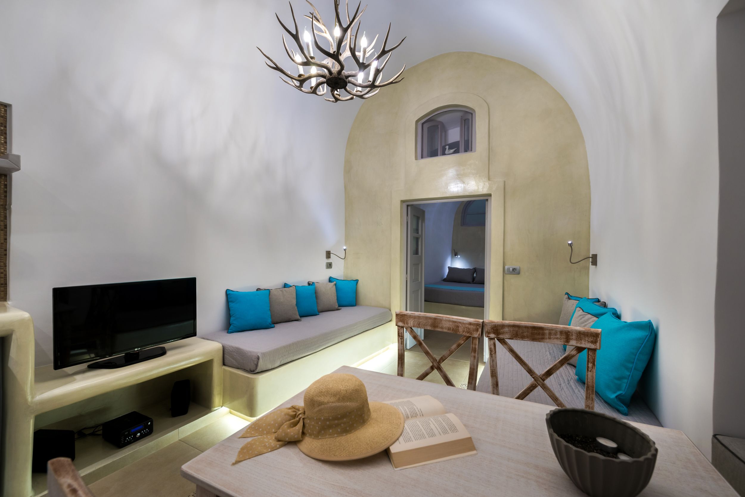 Um quarto aconchegante com televisão de tela plana, um sofá com almofadas azuis, uma mesa de jantar com chapéu e livro, e uma porta em arco peculiar.