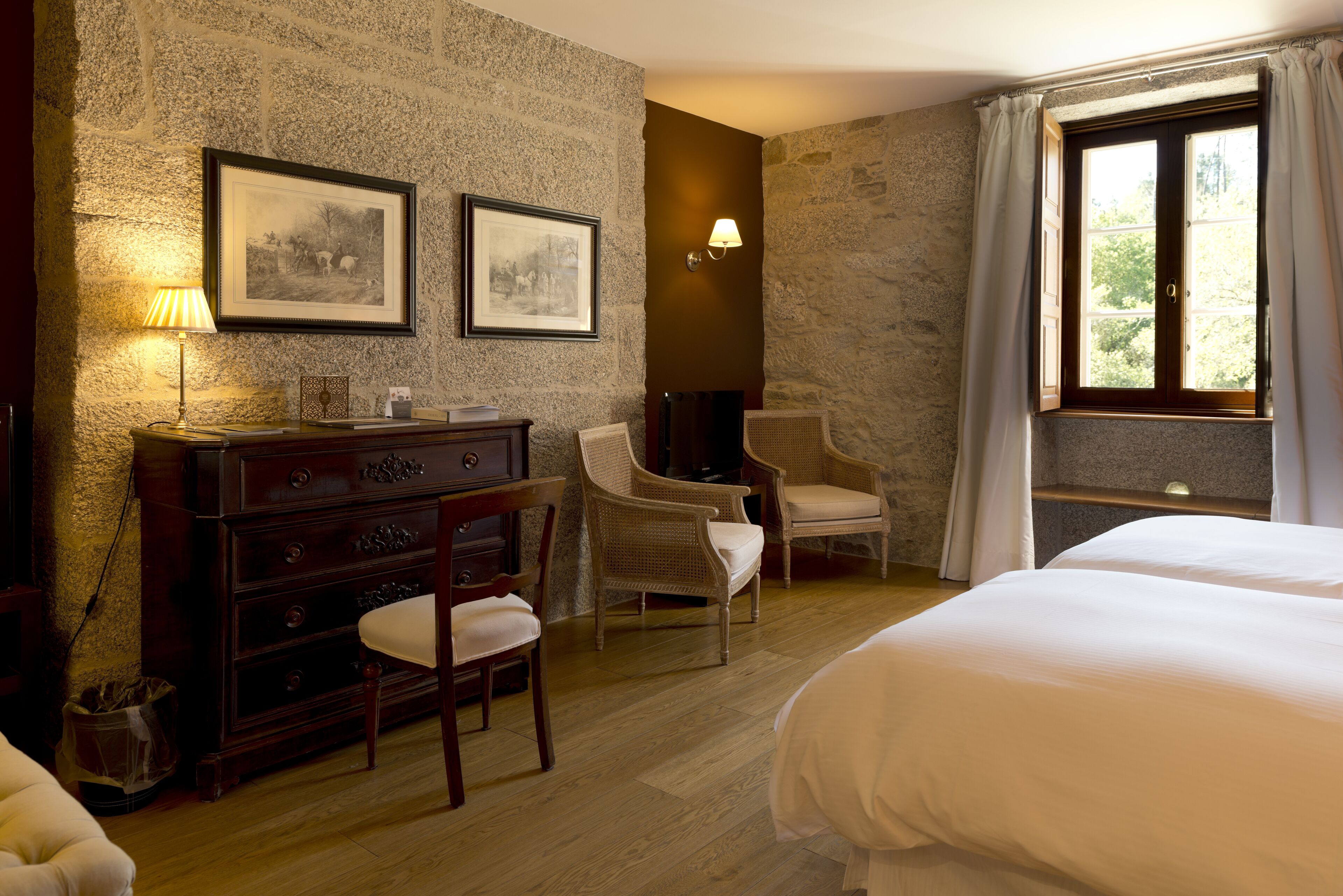 Quarto de hotel com duas camas, penteadeira de madeira, uma cadeira, abajur e duas imagens emolduradas na parede.