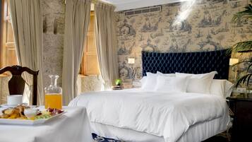 Quarto de hotel com uma cama grande, uma mesa de cabeceira com abajur, bandeja de café da manhã e uma janela com cortinas.