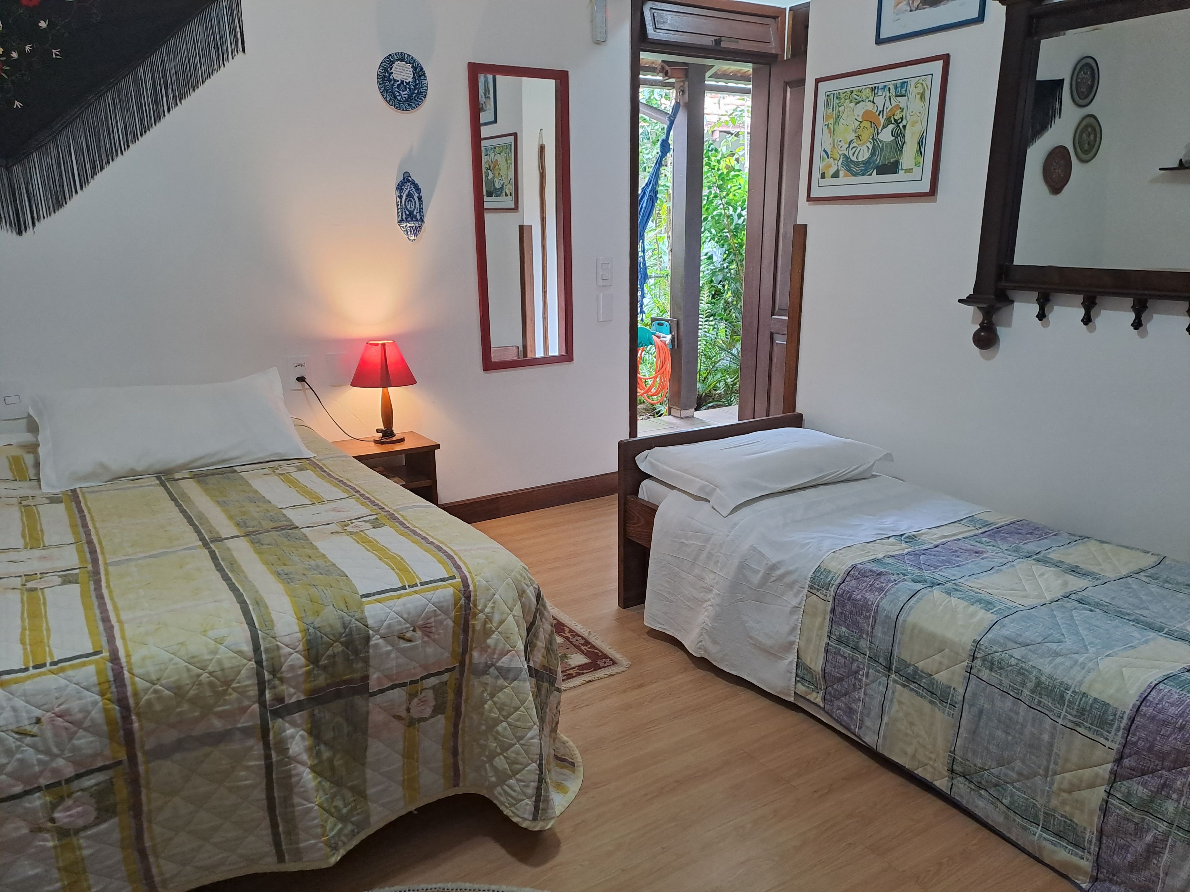 Quarto com duas camas, mesa de cabeceira com abajur, espelho e janela com vista para o verde.
