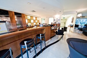 Um saguão de hotel moderno com balcão de recepção, banquetas de bar e uma área de estar com sofá e cadeiras.