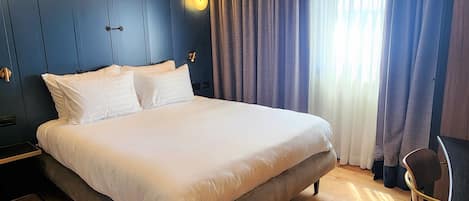 Cama bem arrumada com lençóis brancos, cabeceira azul escura e janela com cortinas.