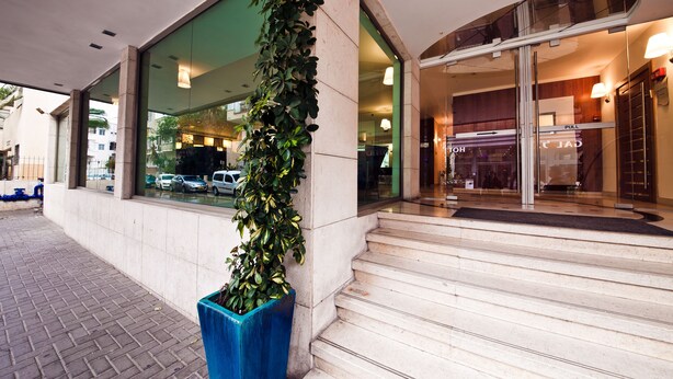 Entrada de hotel com fachada de vidro, escadaria que leva à entrada e um vaso azul com folhagem verde.