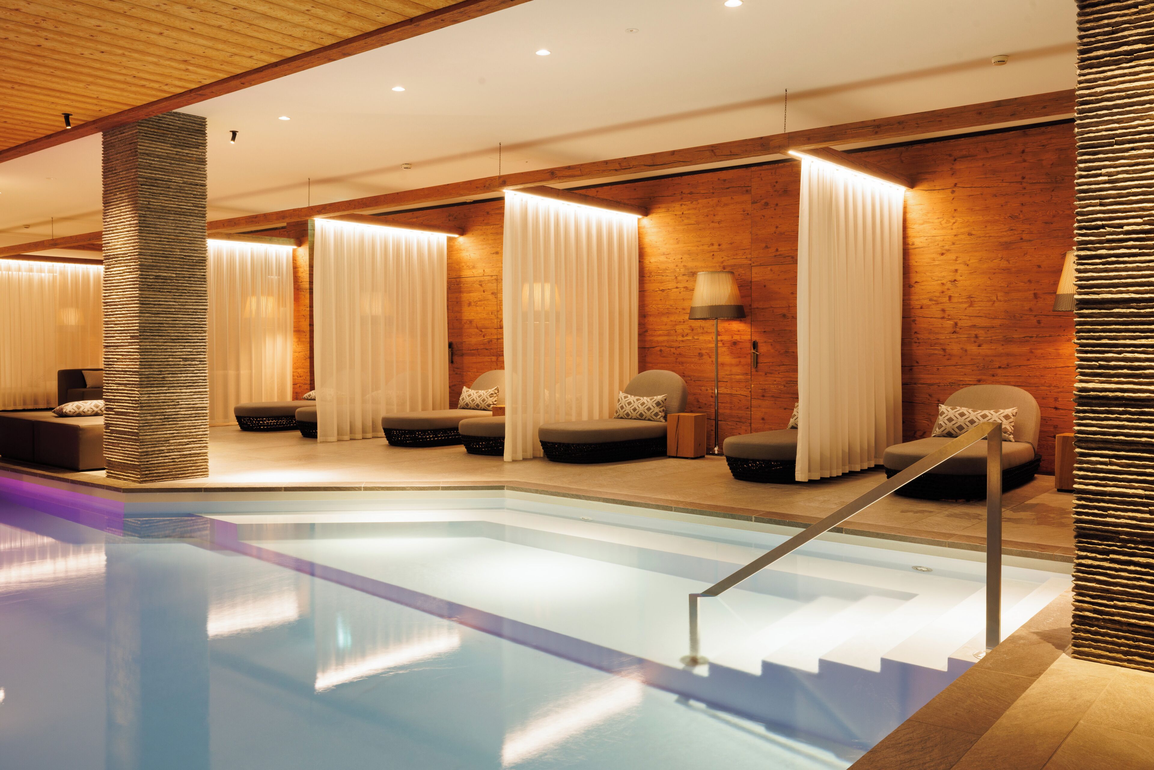 Uma área interna de piscina moderna com piscina transparente, paredes de madeira e áreas de estar com cortinas.