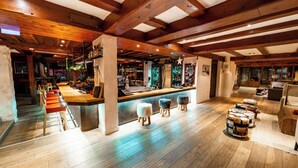 Um espaço amplo de bar com vigas de madeira, balcão, banquetas e uma área de estar com tapete.