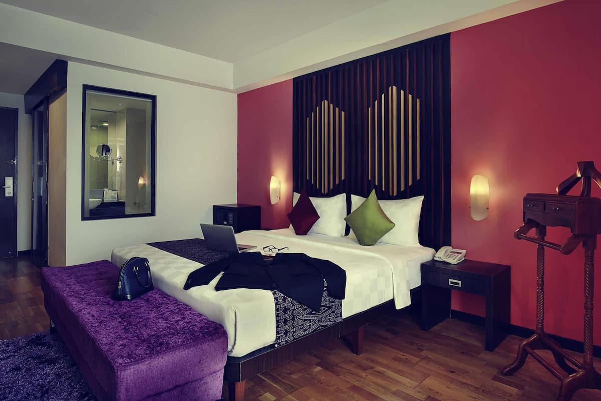 Quarto de hotel moderno com uma cama grande, uma escrivaninha, uma cadeira, um banquinho roxo, um espelho e piso de madeira.