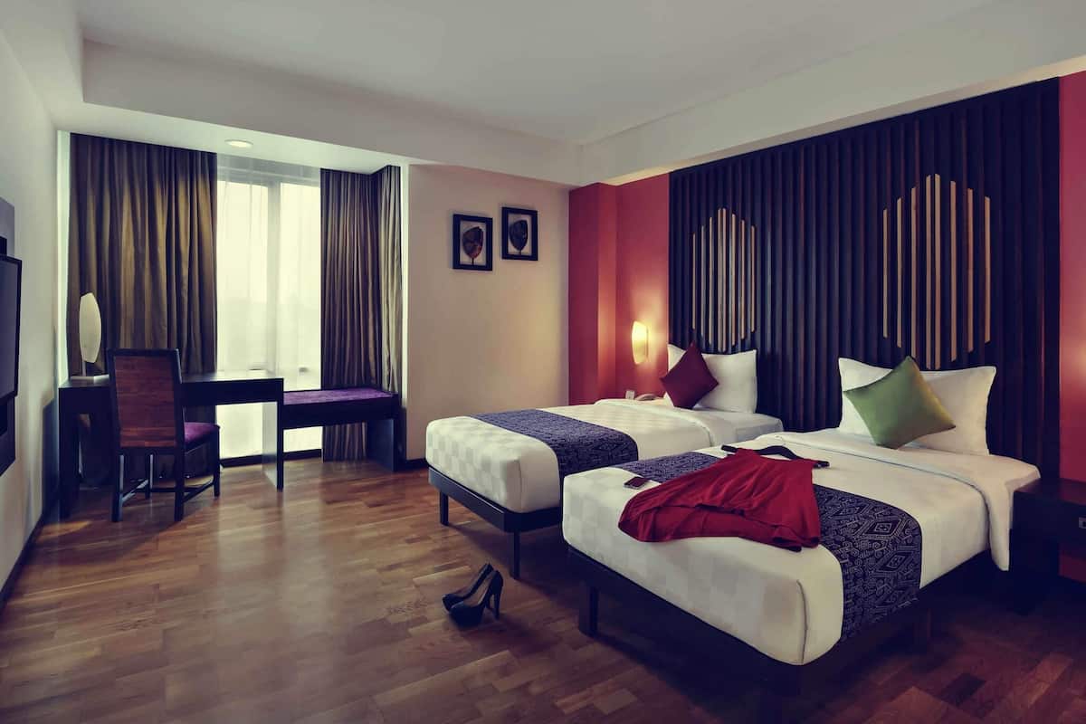 Quarto de hotel com duas camas, uma escrivaninha, uma cadeira e uma janela grande com cortinas.