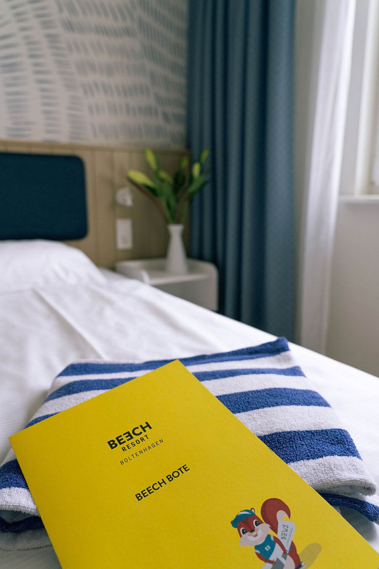 Quarto de hotel com cama bem arrumada, toalha listrada e um livrete amarelo com a inscrição 'BEECH Resort' sobre a cama.
