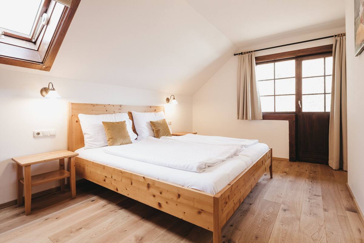 Quarto com cama de madeira, mesa de cabeceira, pequena mesa, janela com cortinas e um claraboia.