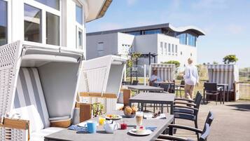 Uma área de refeições ao ar livre com cadeiras e mesas de vime, um buffet de café da manhã com torradas, frutas e bebidas, e um edifício moderno ao fundo.