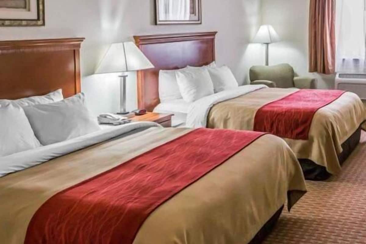 Quarto de hotel com duas camas, cabeceira de madeira, luminária e uma cadeira.