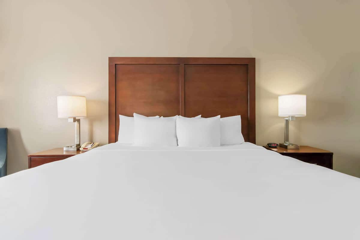Cama bem arrumada com lençóis brancos, cabeceira de madeira e duas mesas de cabeceira com abajures e um telefone.