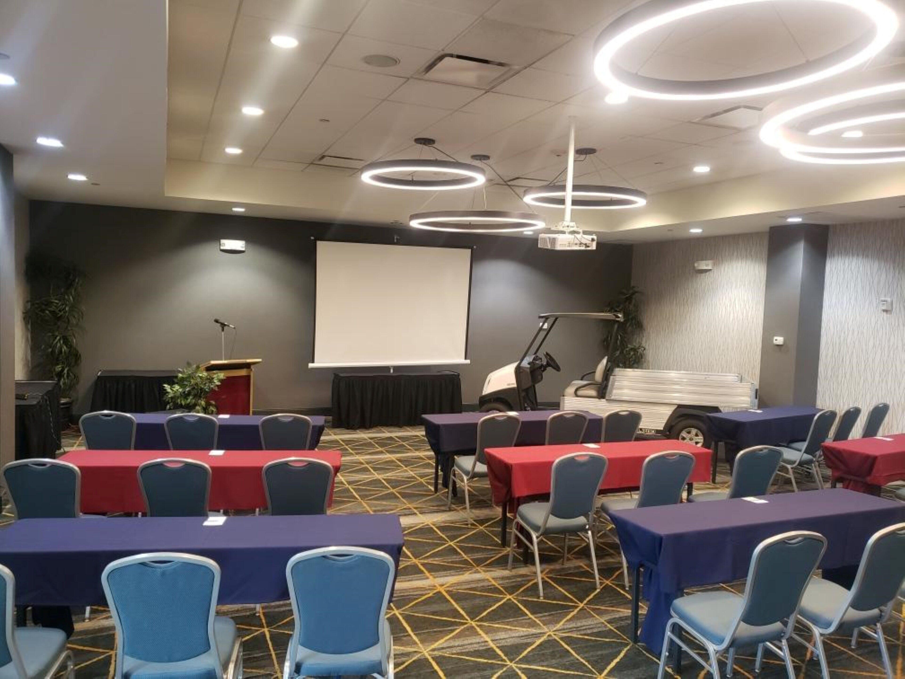 Uma sala de conferências com mesas, cadeiras, um pódio, uma tela de projetor e um carrinho de golfe.