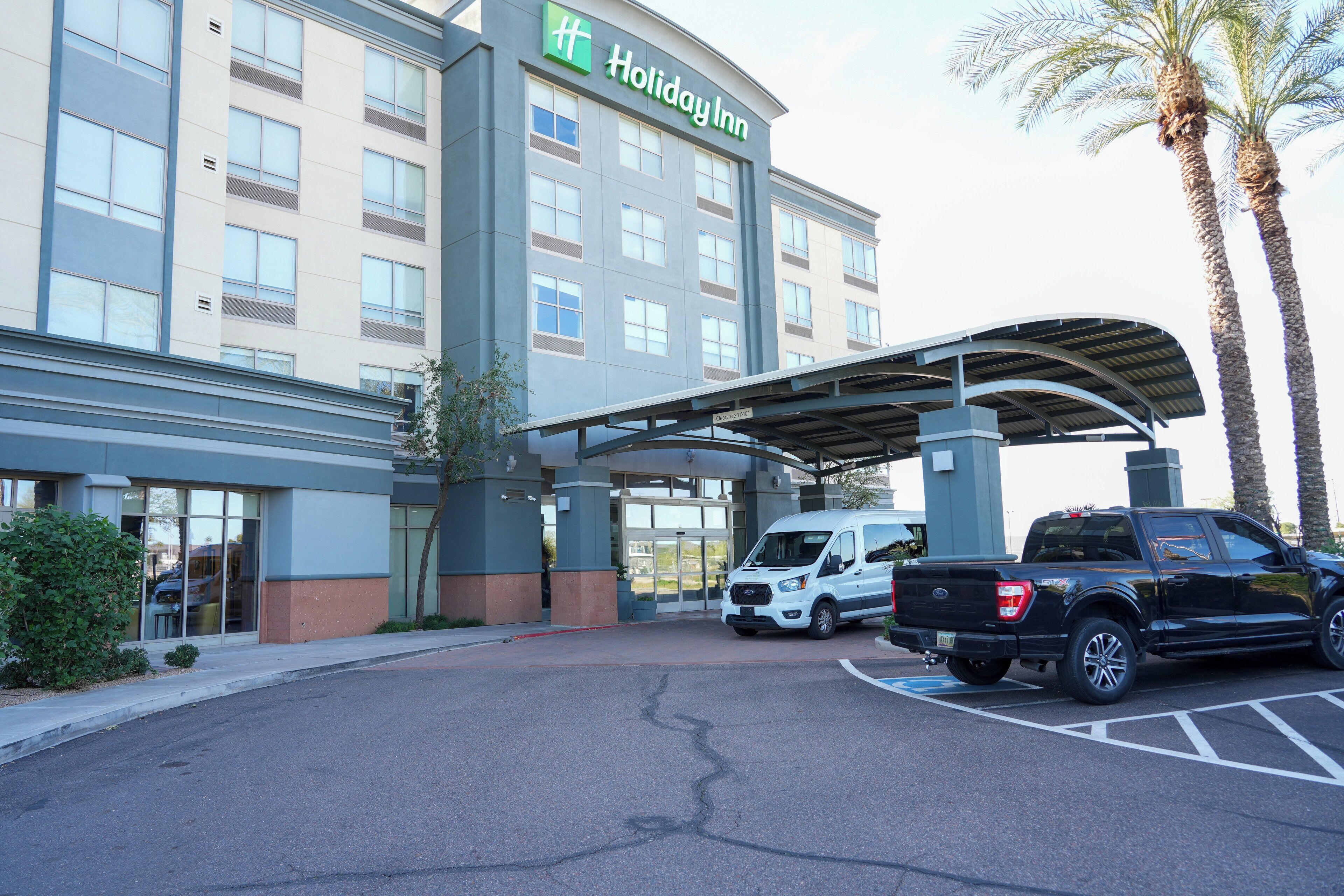 Um hotel Holiday Inn com entrada coberta, estacionamento e uma palmeira ao fundo.