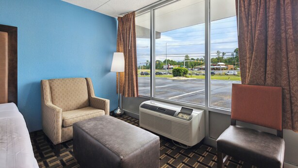 Quarto de hotel com cama, duas poltronas, mesa, luminária e uma janela grande com cortinas.