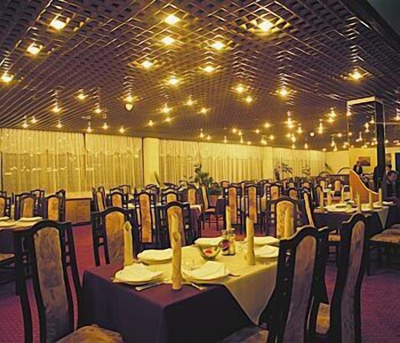 Uma sala de banquetes com fileiras de mesas preparadas para refeições, teto alto com padrão geométrico e área de palco ao fundo.