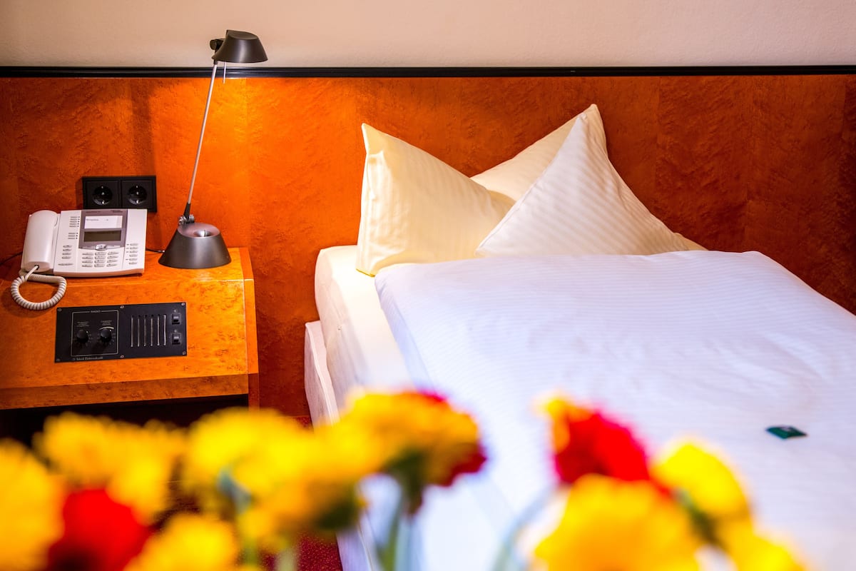 Quarto de hotel com cama, mesa de cabeceira com telefone e luminária, e parede com acabamento texturizado em laranja.