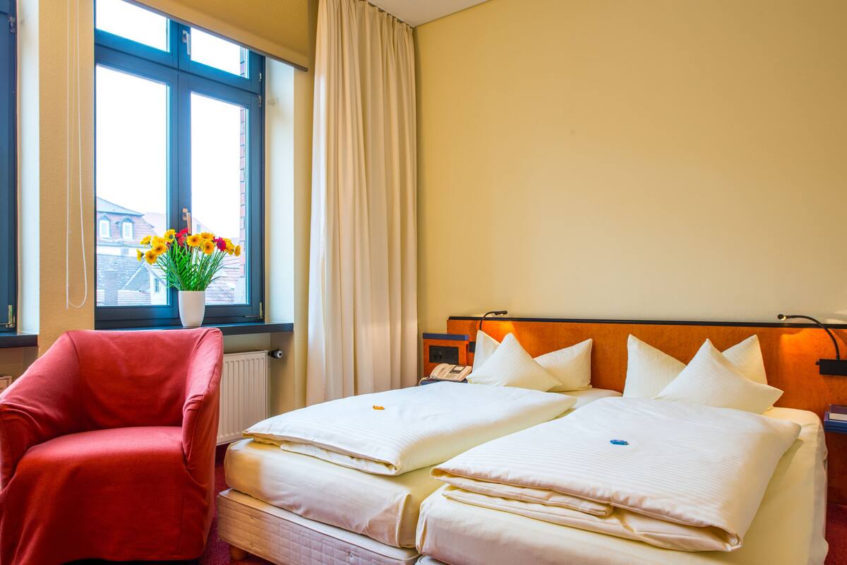 Quarto de hotel com cama, uma cadeira, janela com cortinas e vista para edifícios.