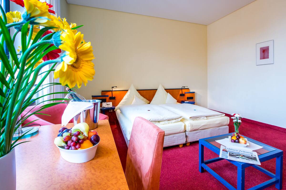 Quarto de hotel com uma cama, uma mesa de cabeceira com um jornal, uma fruteira e um vaso de flores.