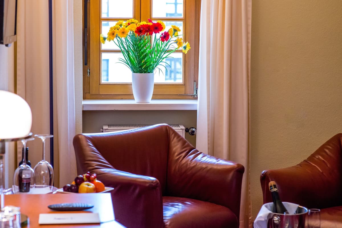 Quarto com poltrona de couro, mesa com uma garrafa de champanhe, um vaso de flores e janela com cortinas.