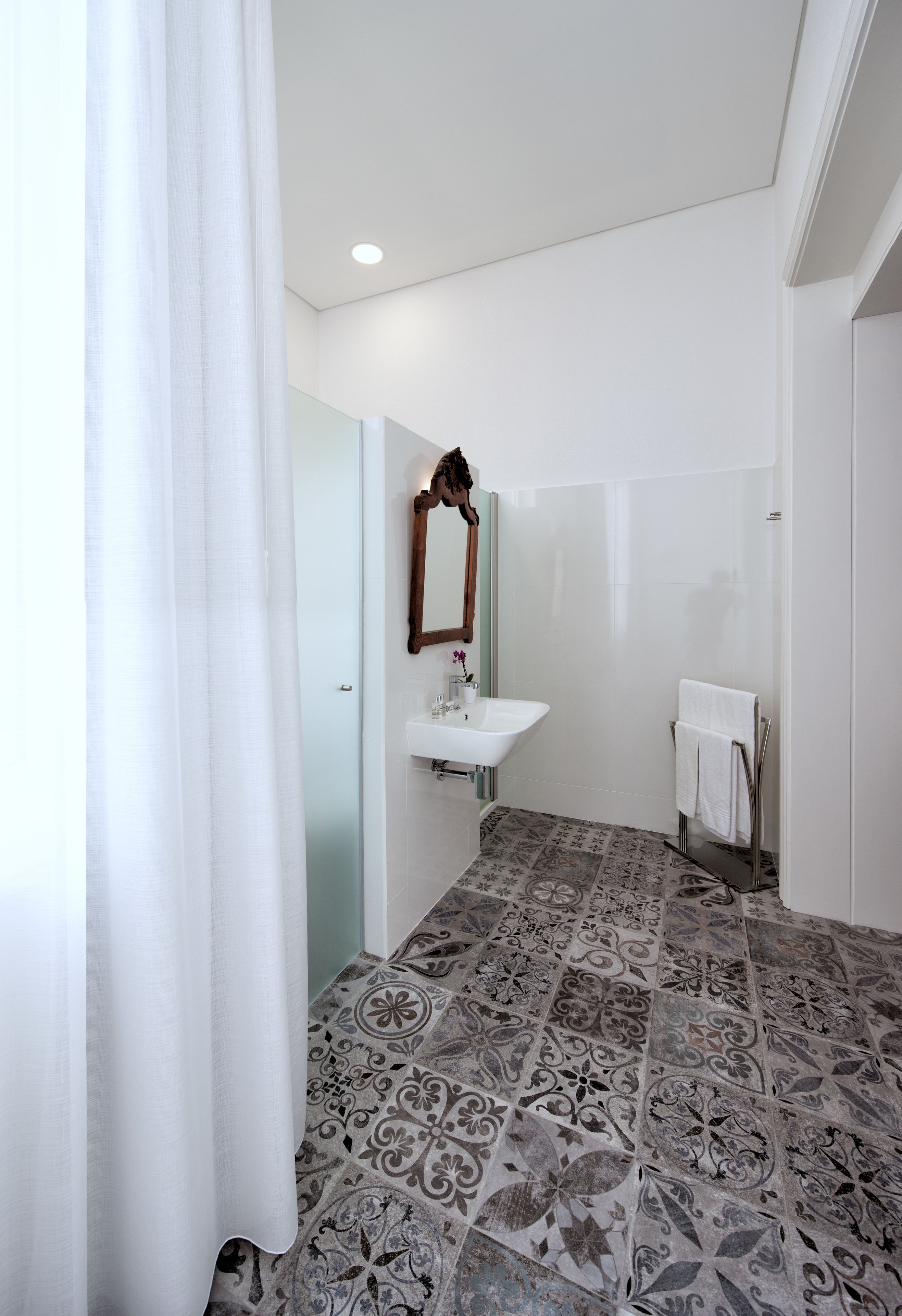 Um banheiro com piso de azulejos estampados, pia branca, espelho e porta-toalhas.