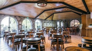 Um espaço interno amplo com mesas e cadeiras de madeira, paredes de pedra e grandes janelas em arco que permitem a entrada de luz natural.