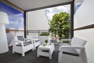 Uma varanda com móveis de vime branco, uma mesa de vidro e vista para uma árvore e um edifício.