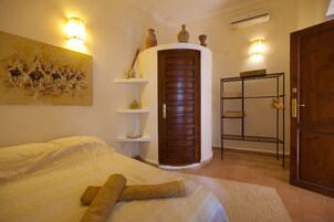 Quarto com cama, porta de madeira, prateleira com objetos decorativos e luminária fixada na parede.