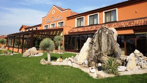 Hotel com um amplo jardim de pedras na frente, área de varanda coberta e um céu azul límpido.
