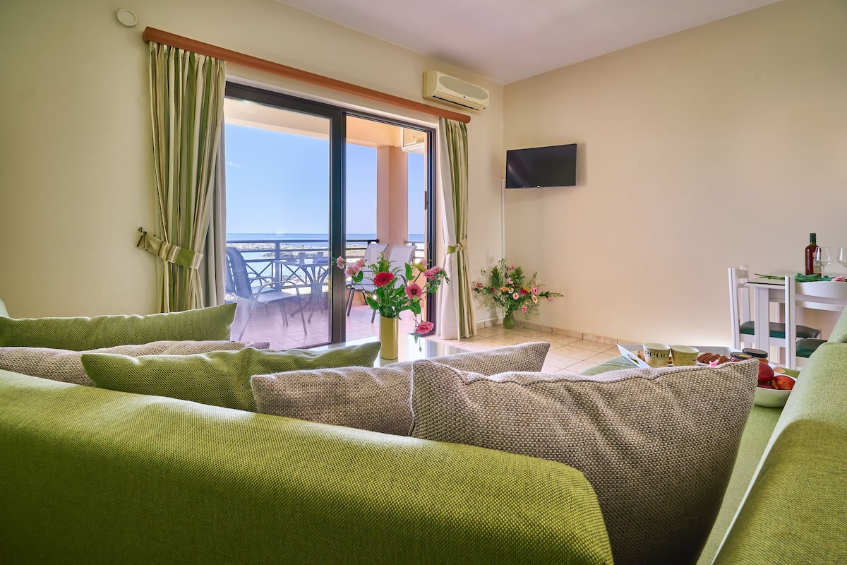 Sala de estar com sofás verdes, uma televisão de tela plana, uma mesa de jantar com garrafa e copos, e uma varanda com vista.