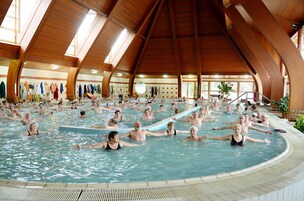 Uma piscina coberta ampla, com várias pistas de natação, vigas de madeira e teto alto.