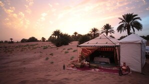 Uma tenda no deserto com uma cama e uma mesa pequena, rodeada por palmeiras e dunas de areia.