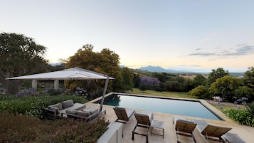 Uma área de piscina com espreguiçadeiras e um pérgola, cercada por vegetação exuberante e vista para montanhas distantes.